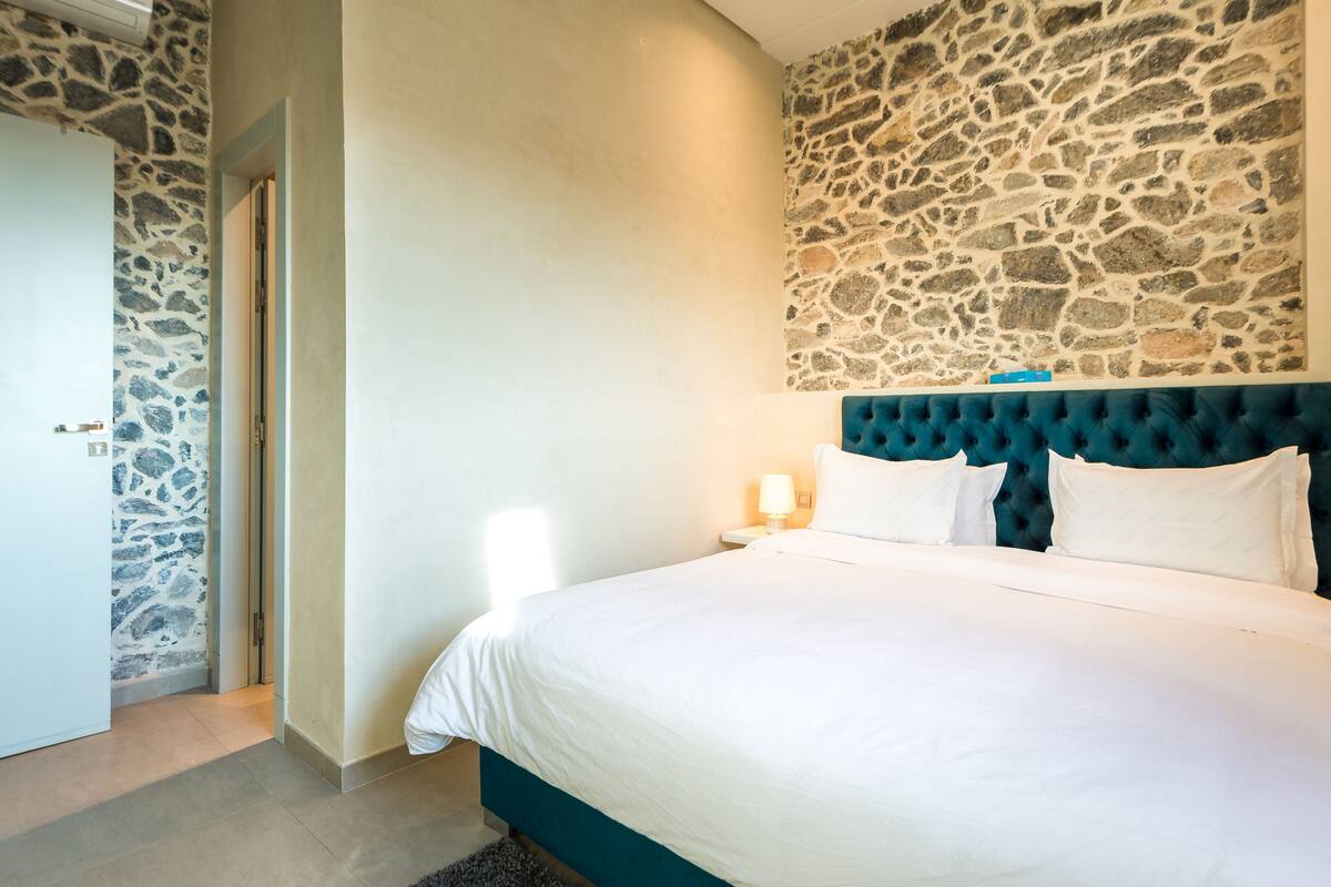 Quarto com parede de pedra, cabeceira estofada em tons de verde-água e colcha branca.