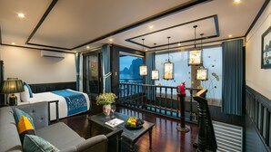 Quarto de hotel com cama, sofá, mesa de jantar com frutas, um vaso de flores e vista para o mar através de grandes janelas.