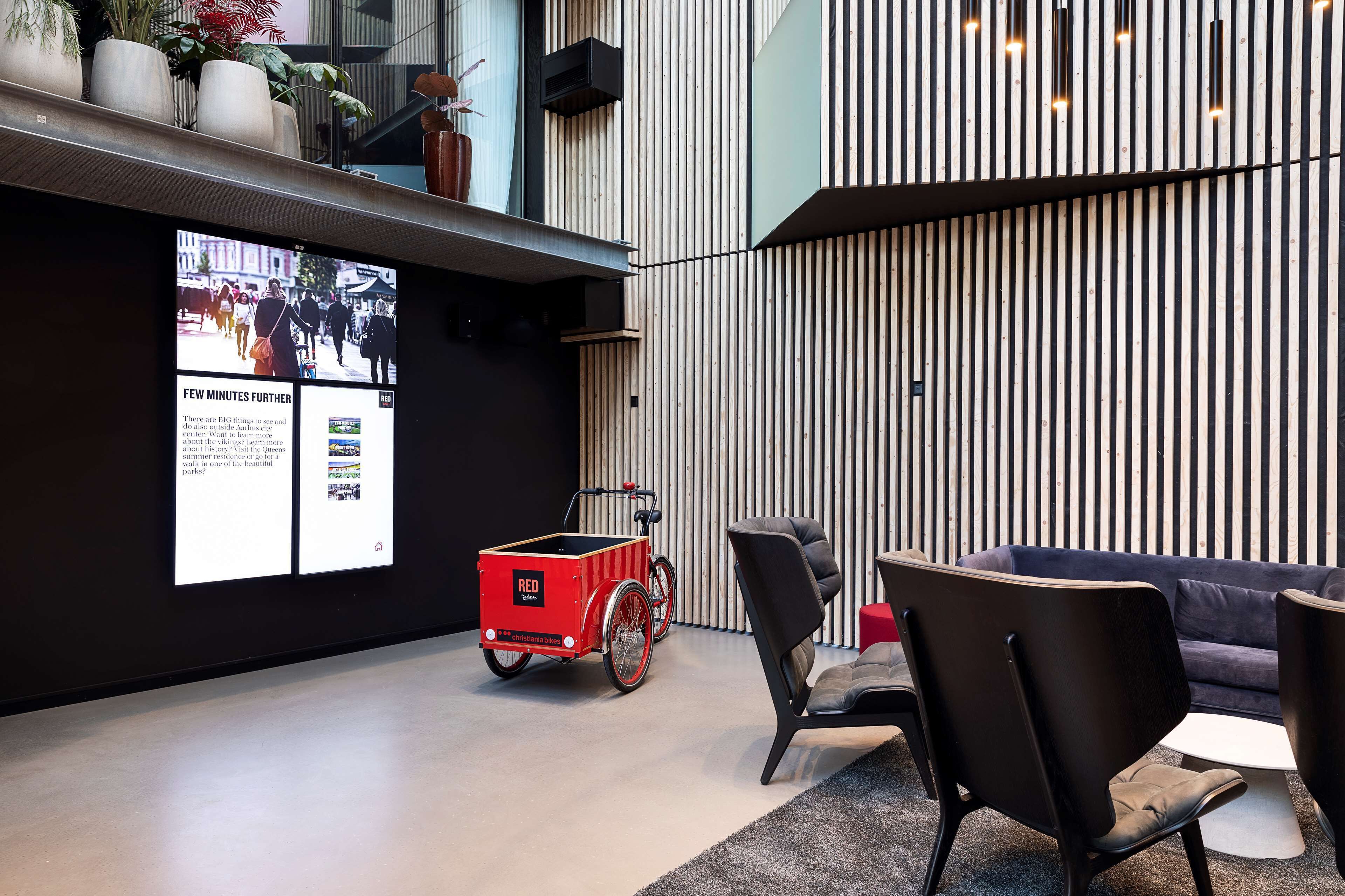 Um interior moderno com um painel digital, uma bicicleta de entrega vermelha e mobiliário contemporâneo.