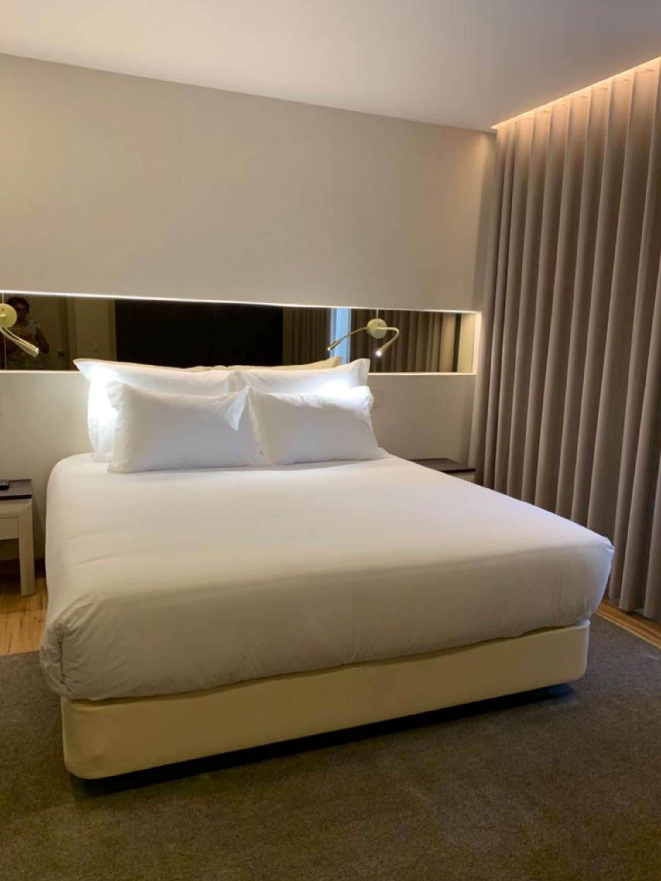 Quarto de hotel moderno com uma cama grande, mesa de cabeceira, luminária e janela com cortinas.