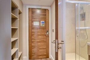 Quarto de hotel com porta de madeira, box de chuveiro em vidro e parede com azulejos brancos.