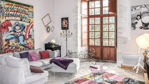 Uma sala de estar com um sofá branco, um tapete colorido, um grande gibi do Capitão América, uma porta de madeira e um candelabro.