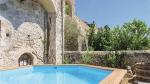 Piscina com paredes de pedra e deck de madeira, cercada por vegetação e um muro de pedra.