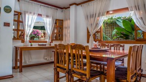 Uma área de refeições com mobiliário de madeira, cortinas brancas e vista para o verde lá fora.
