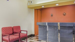 Uma área de recepção com balcão de check-in, duas cadeiras vermelhas estampadas e piso ladrilhado.