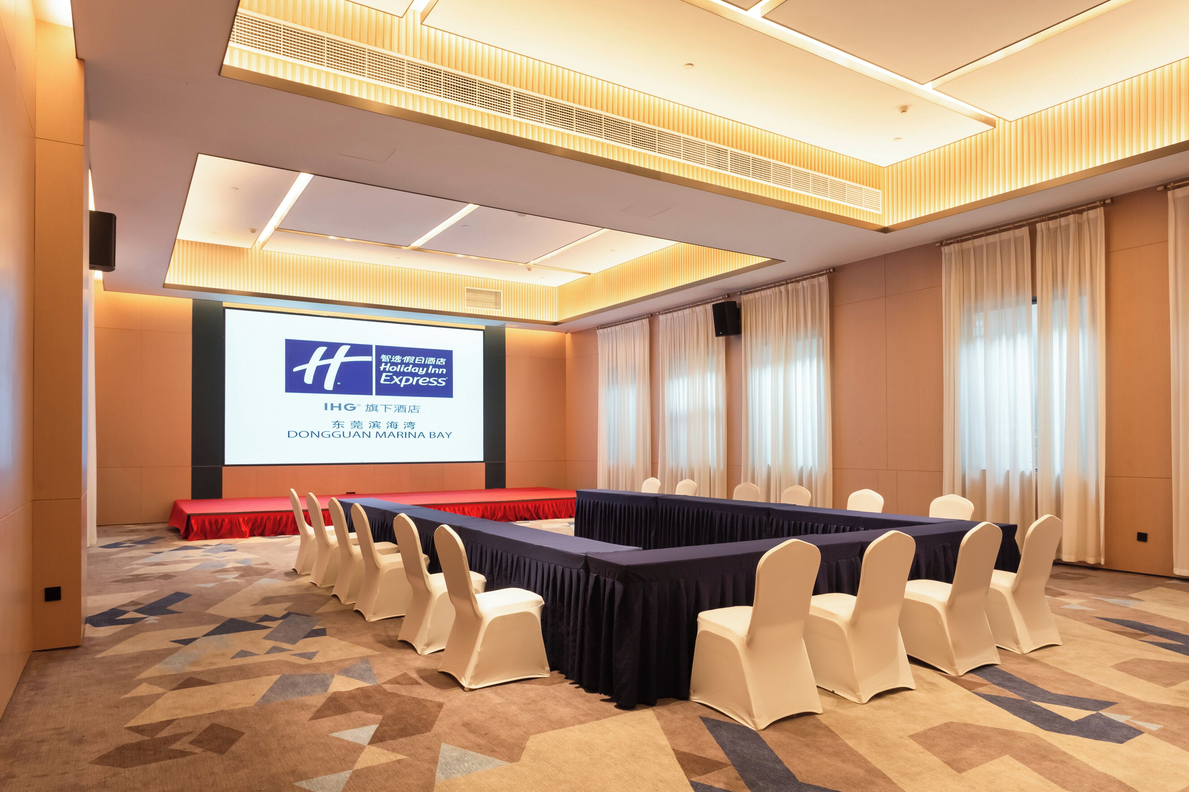 Uma sala de conferências com uma mesa comprida, cadeiras brancas, um palco vermelho e uma tela exibindo informações do hotel.