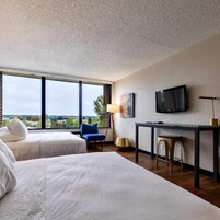 Quarto de hotel com duas camas, uma escrivaninha, uma cadeira, televisão e vista para o exterior.
