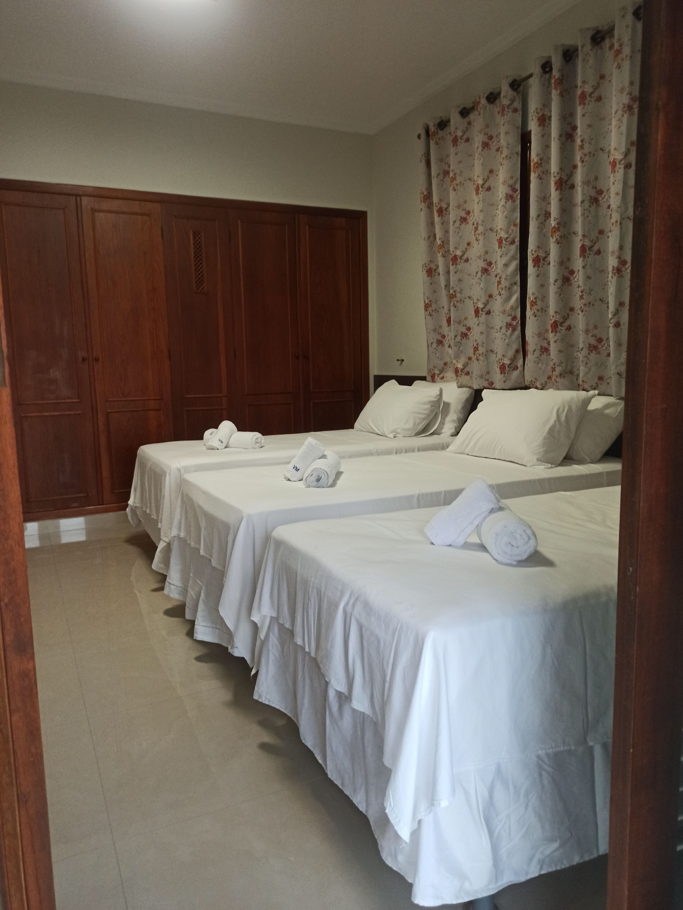 Quarto de hotel com três camas de solteiro, armários de madeira e cortina florida.