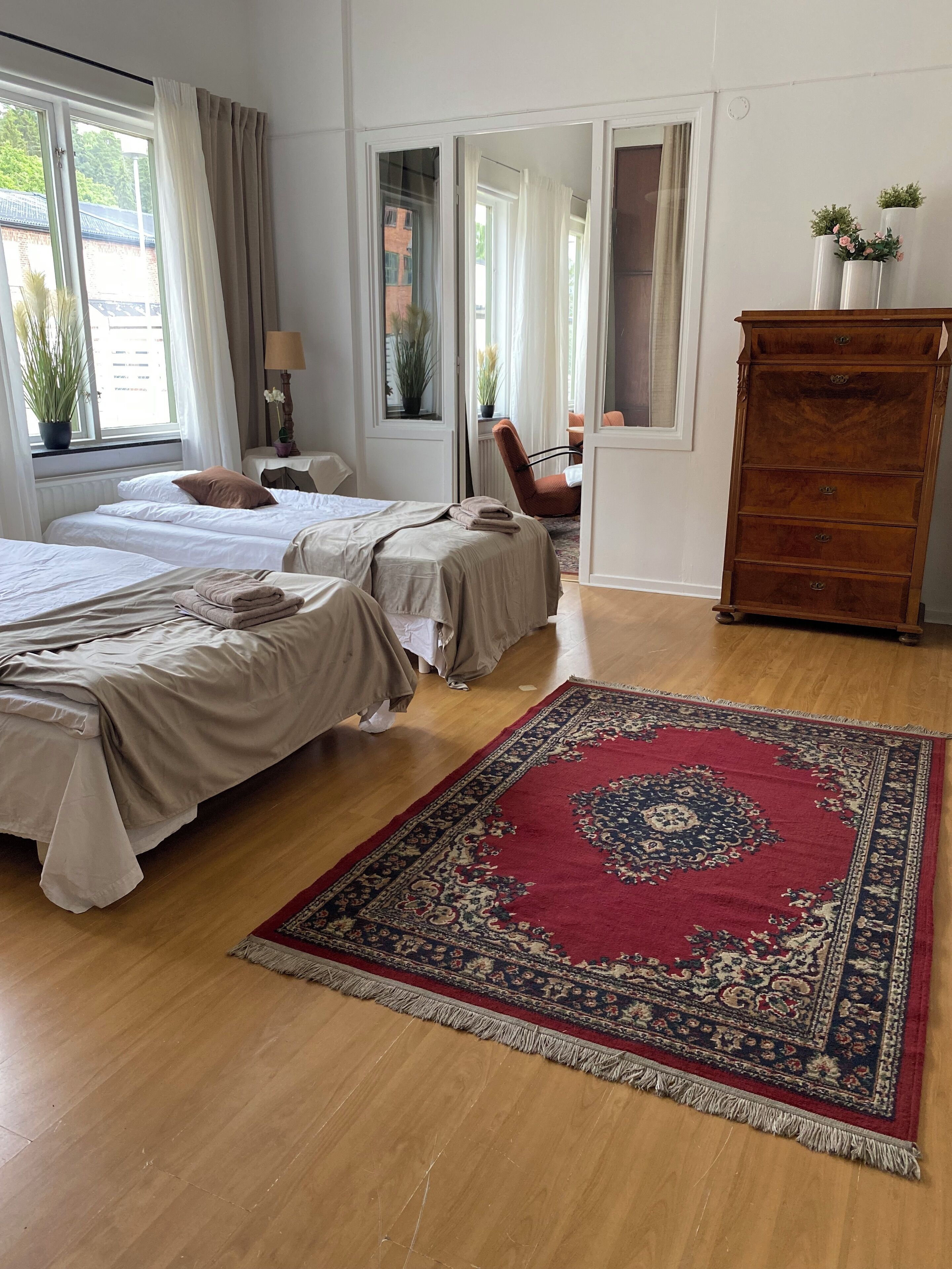 Quarto de hotel com duas camas, cómoda de madeira, tapete vermelho estampado e vista para um edifício pela janela.