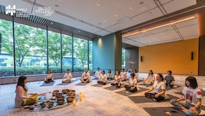 Um grupo de pessoas sentadas em tapetes de yoga em um espaço amplo com grandes janelas, dispostas em círculo, voltadas para uma área central com tigelas e velas.
