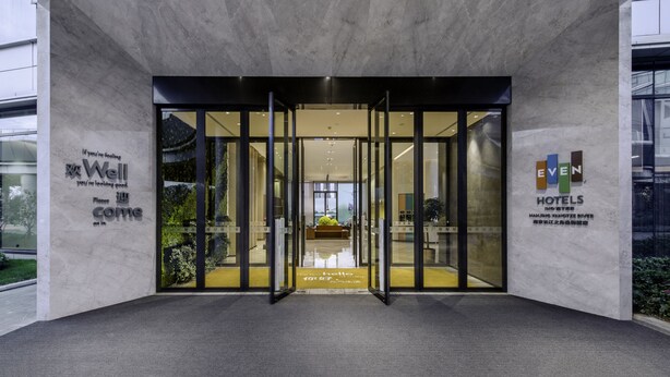 Entrada de um hotel com porta de vidro, uma placa com a inscrição 'Parece que você está ótimo' e o logotipo da marca 'EVEN Hotels'.