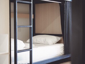 Uma cama de solteiro com colchão e almofadas, uma janela com cortinas e uma tomada na parede.