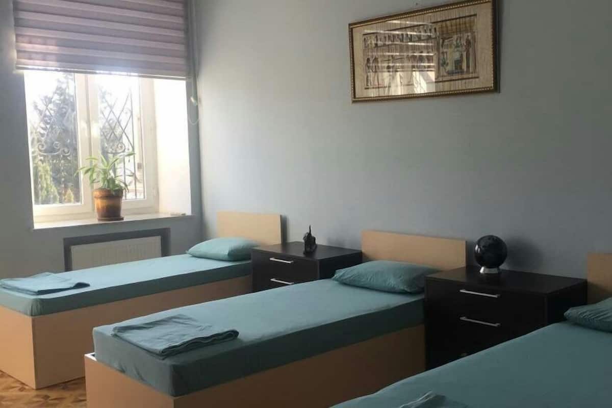 Quarto com três camas simples, janela com vista para áreas verdes, radiador e quadro pendurado na parede.