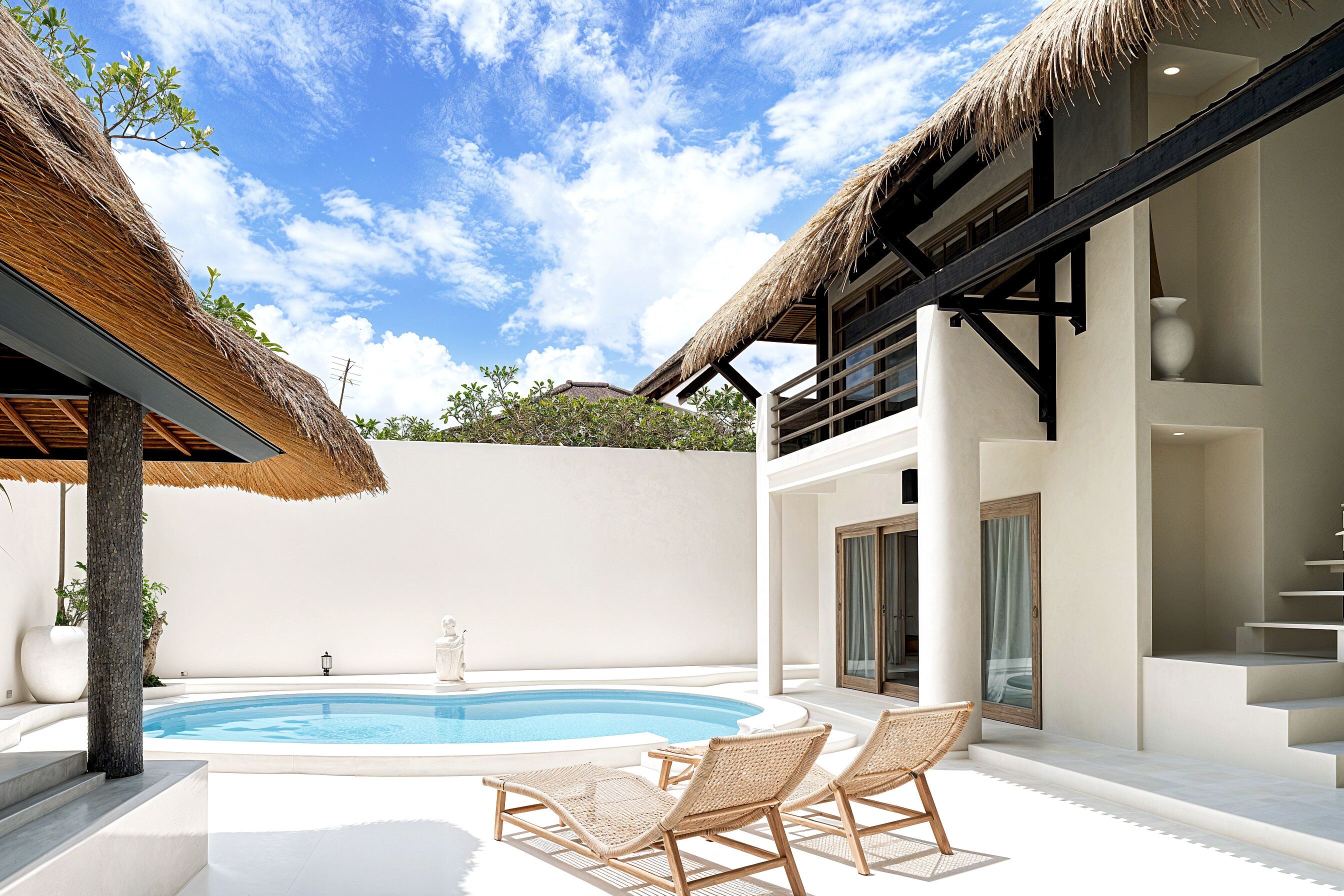 Uma área moderna com piscina ao ar livre, um telhado de palha, duas poltronas de vime e uma escadaria para um andar superior.