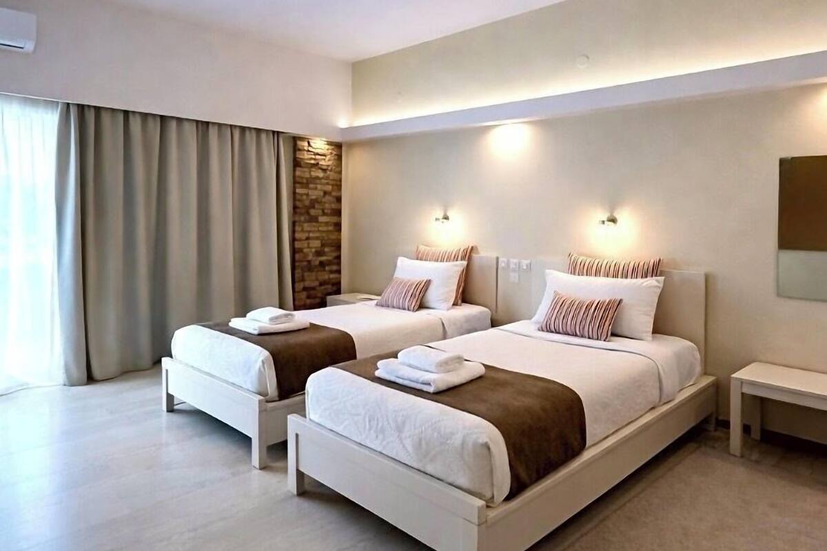 Quarto de hotel moderno com duas camas de solteiro, parede com detalhes em pedra e uma janela grande com cortinas.