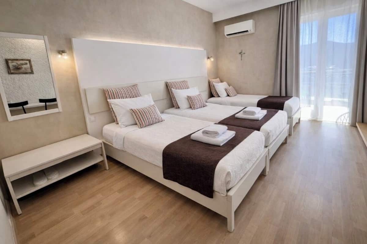 Quarto de hotel com duas camas, um banco e uma janela grande.
