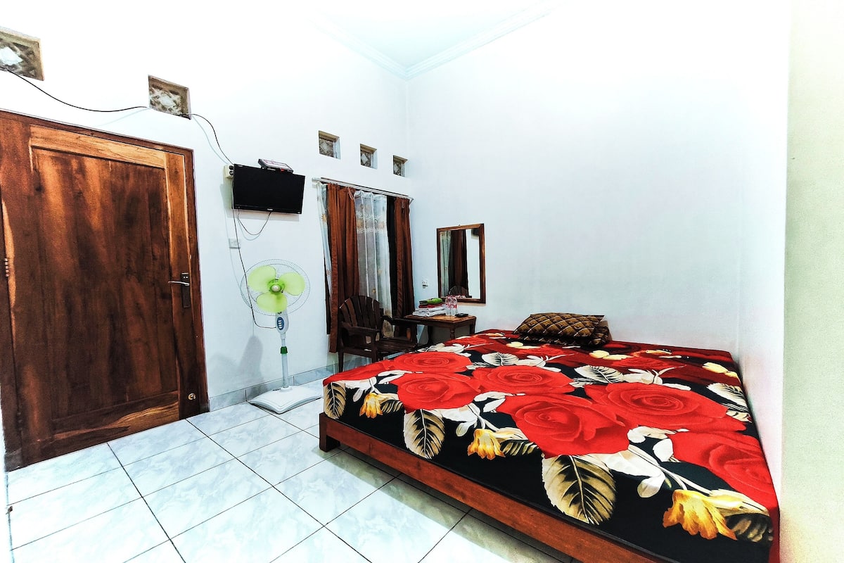 Um quarto com cama, porta de madeira, televisão fixada na parede, uma mesinha e uma cadeira.