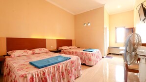 Quarto de hotel com duas camas, cada uma com roupa de cama floral e um cobertor azul. Há um ventilador portátil e uma televisão fixada na parede.