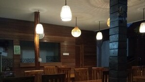 Interior de restaurante com mesas e cadeiras de madeira, luminárias pendentes e um pilar de pedra.