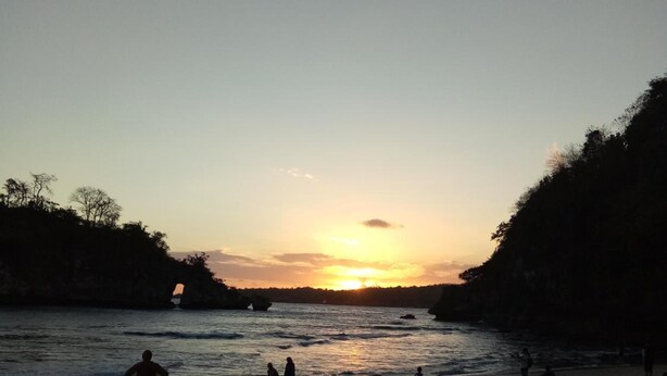 Cena de praia ao pôr do sol com pessoas caminhando e sentadas na areia.