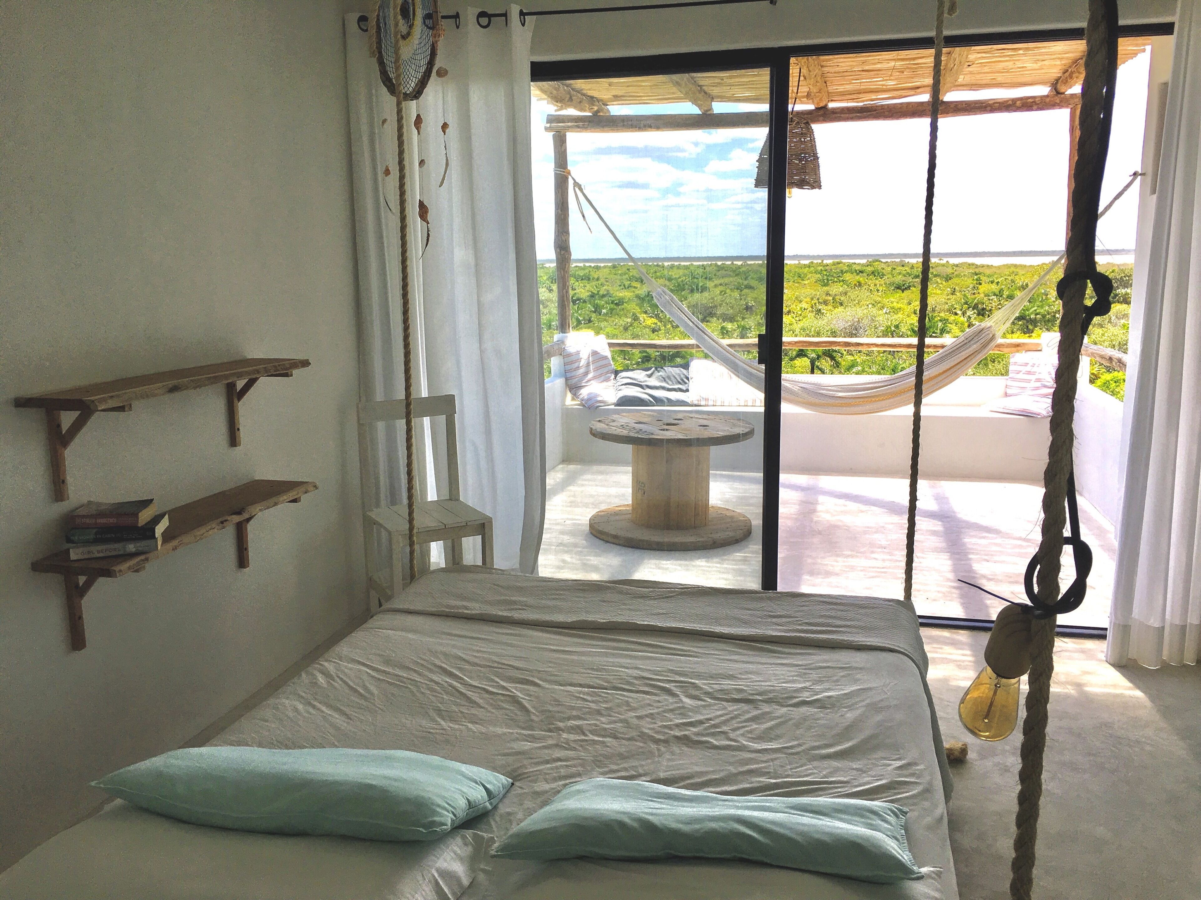 Quarto com cama, almofadas, uma cadeira, uma mesinha e uma rede lá fora da janela.
