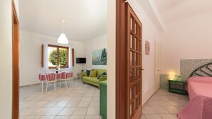 Quarto com área de jantar, um sofá verde, cama com colcha rosa e televisão na parede.