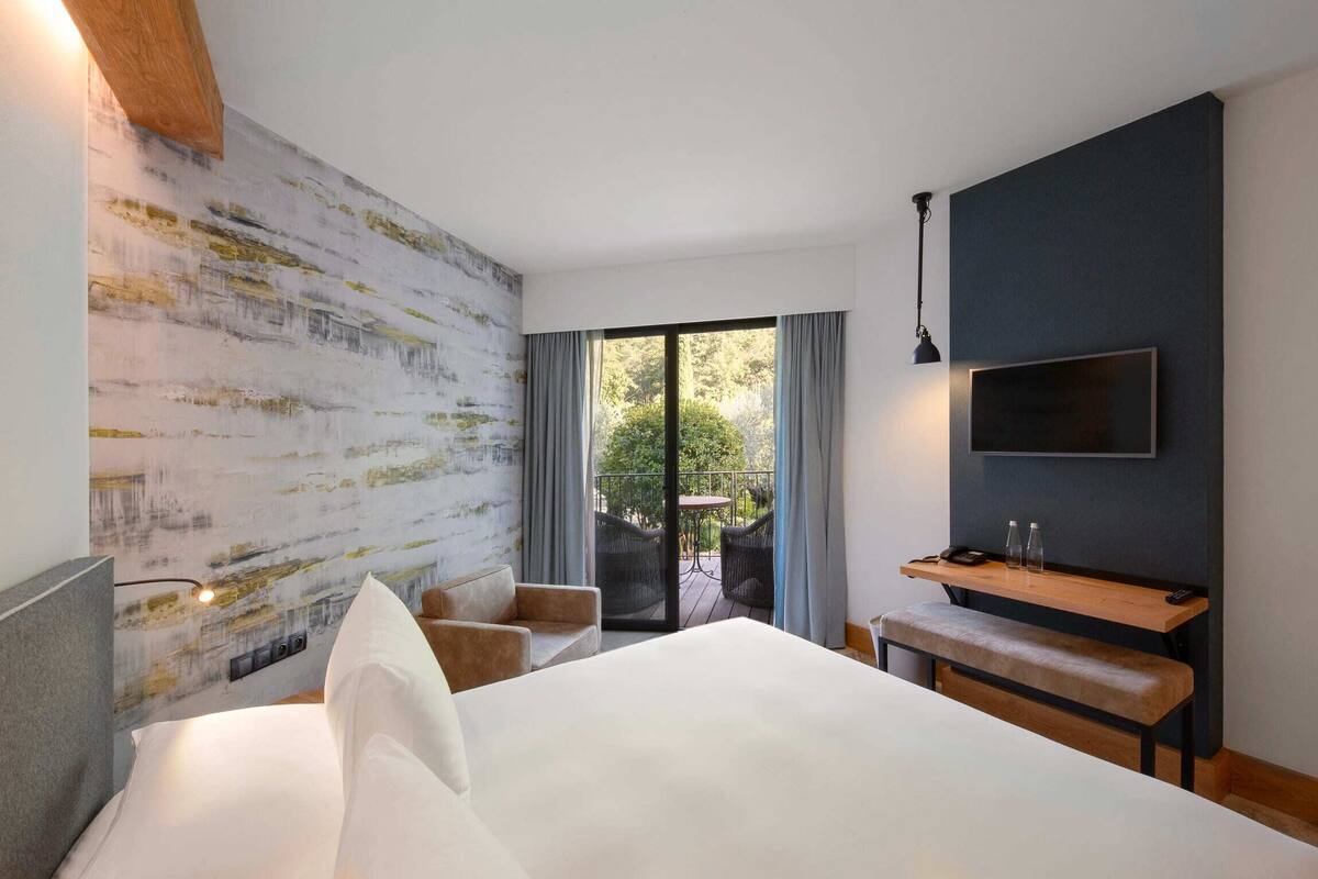 Quarto de hotel moderno com uma cama grande, televisão de tela plana na parede, uma mesa pequena com um banco e uma varanda com área de estar externa.