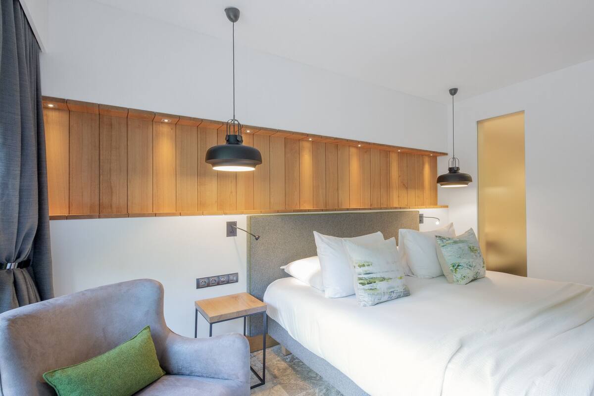 Quarto de hotel moderno com uma cama, uma cadeira, uma mesinha e painéis de madeira na parede.
