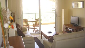 Uma sala de estar com sofá, mesa de centro, televisão em um móvel, varanda com cadeiras e um vaso com flores.