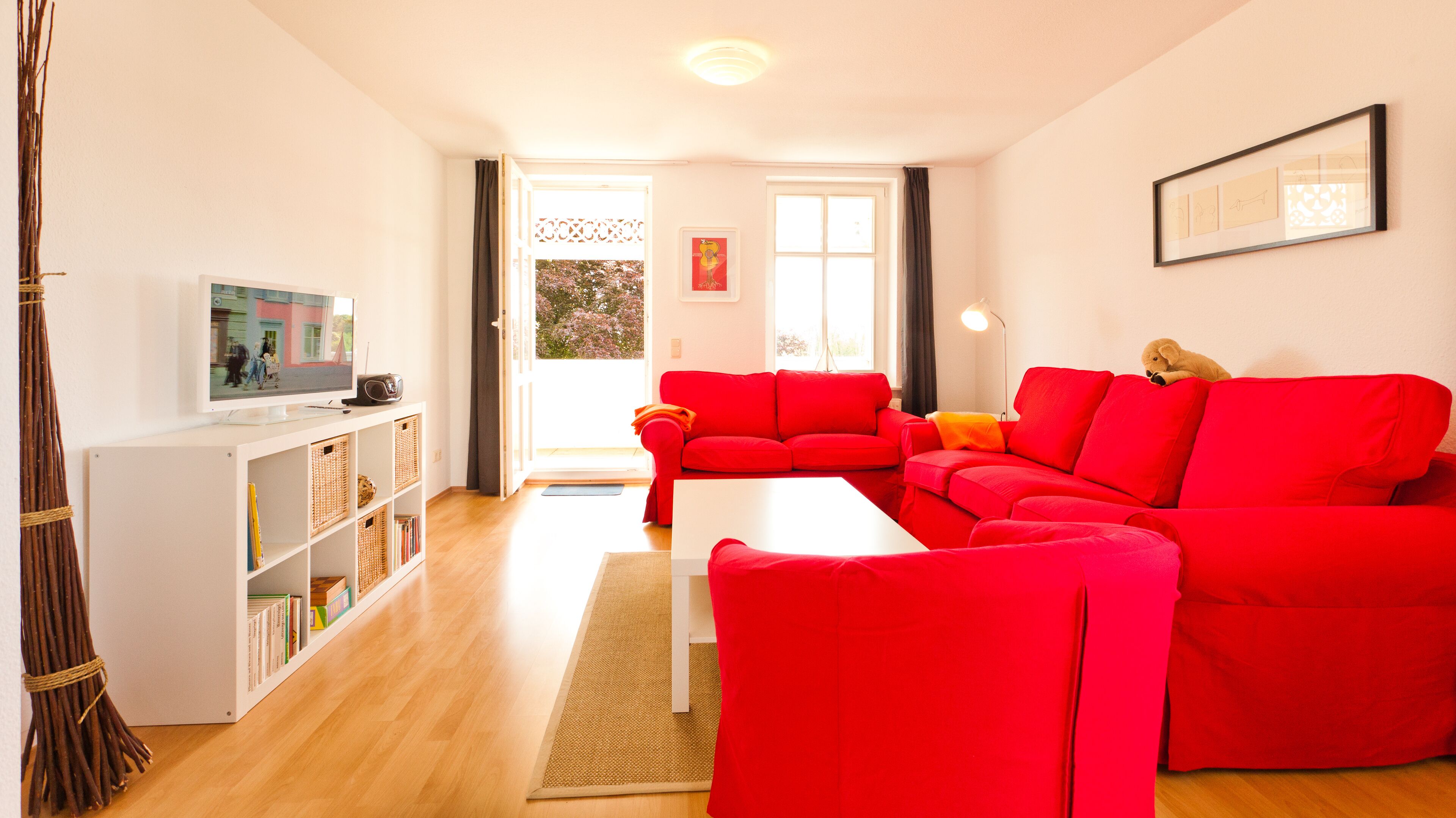 Uma sala de estar luminosa com um sofá vermelho, uma mesa de centro branca e uma televisão sobre um suporte branco.