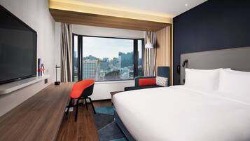 Quarto de hotel com uma cama grande, uma escrivaninha, uma cadeira, uma televisão e uma janela com vista para a cidade.