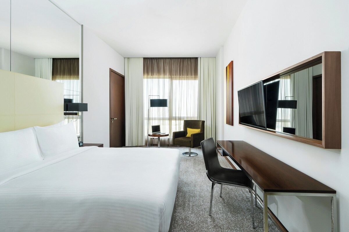 Quarto de hotel com uma cama grande, uma escrivaninha, uma cadeira, uma televisão e uma janela com cortinas.