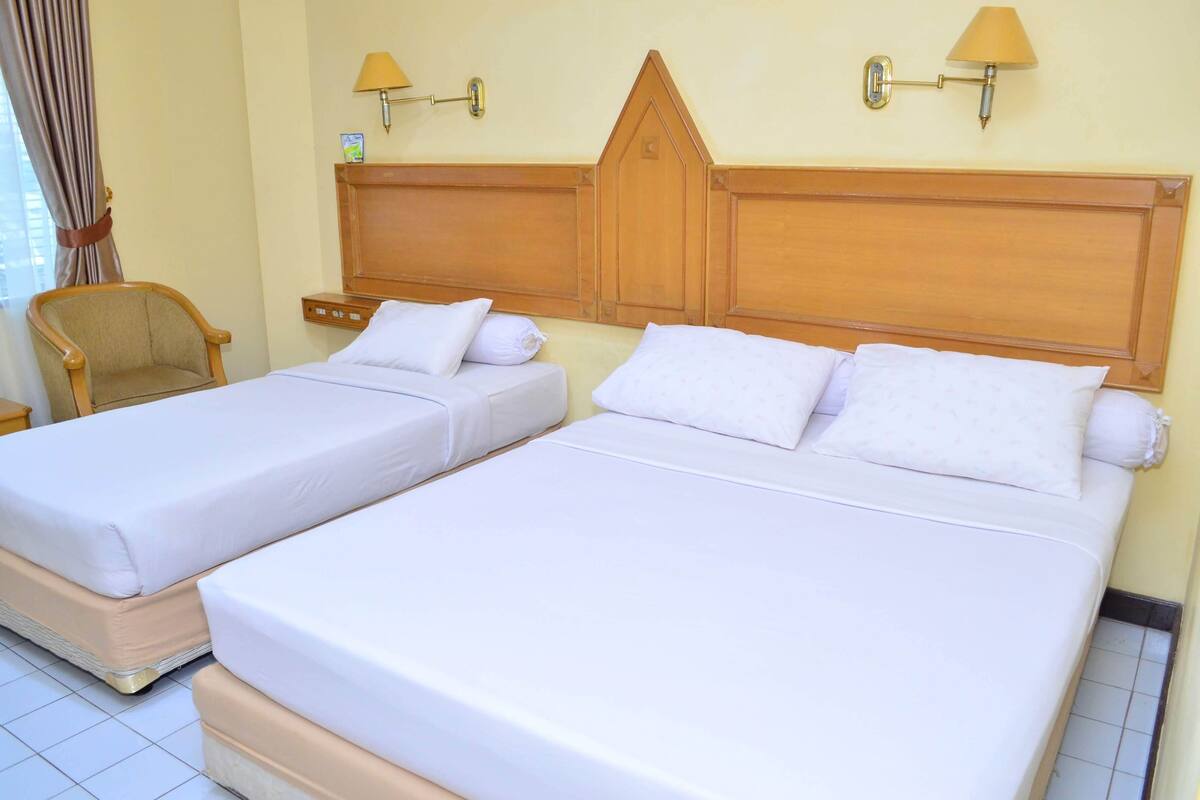 Quarto de hotel com duas camas de solteiro, cabeceira de madeira, lumin árias de cabeceira, uma cadeira e uma pequena mesa.