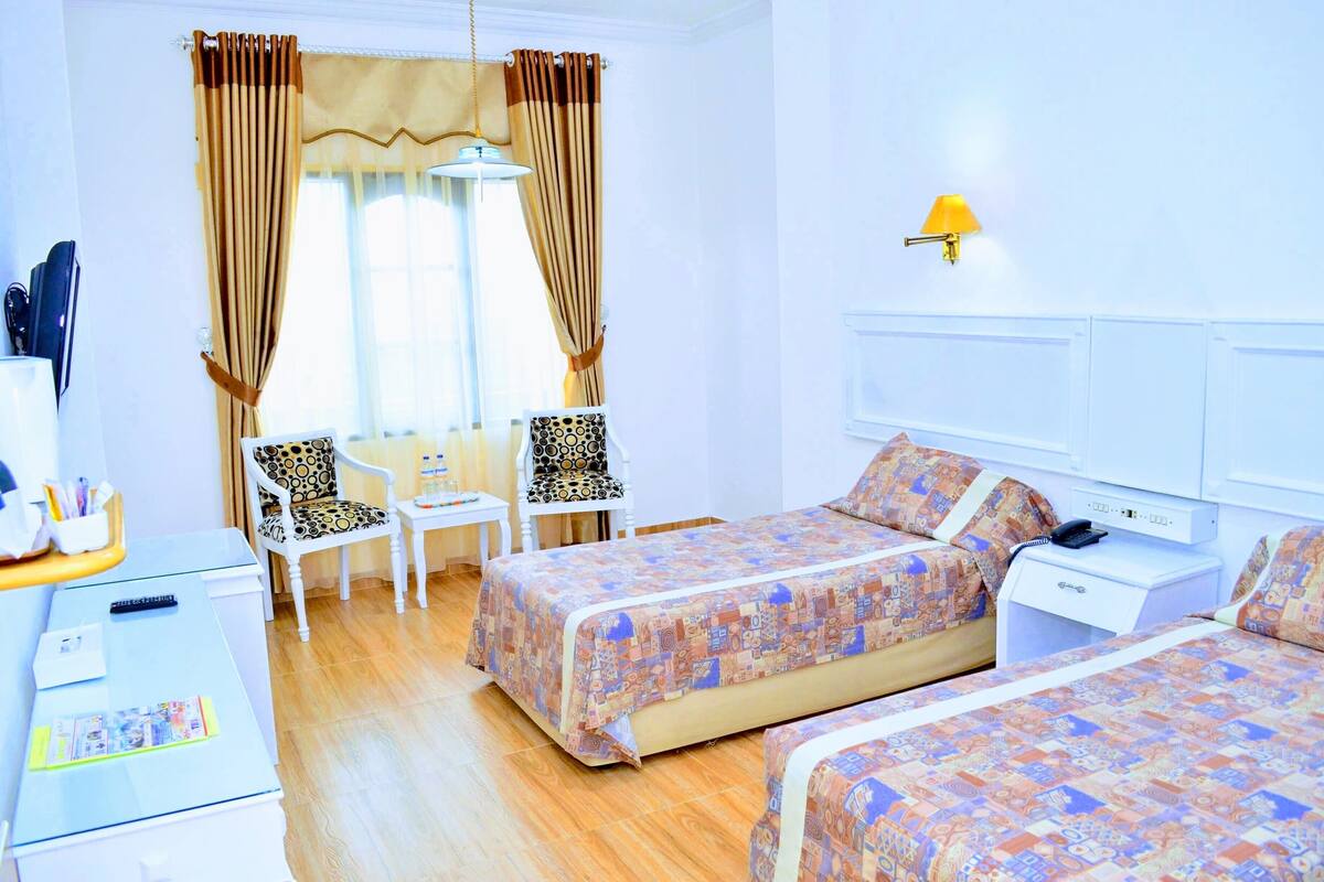 Quarto de hotel com duas camas, uma escrivaninha, cadeiras, luminária e janela com cortinas.