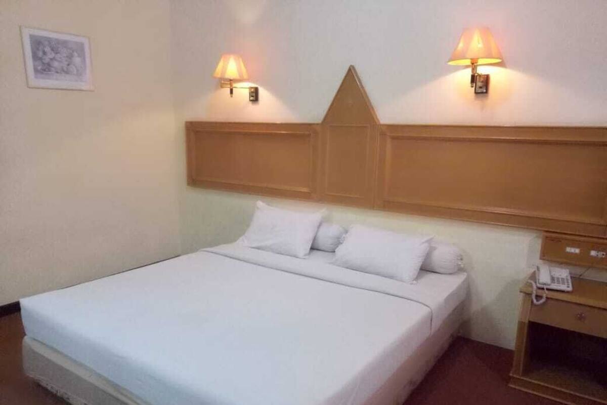 Quarto de hotel com uma cama de solteiro, duas luminárias fixadas na parede, uma mesa de cabeceira com telefone e um quadro na parede.