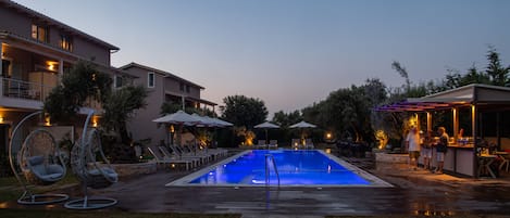 Piscina com espreguiçadeiras e guarda-sóis, cercada por edifícios e árvores.