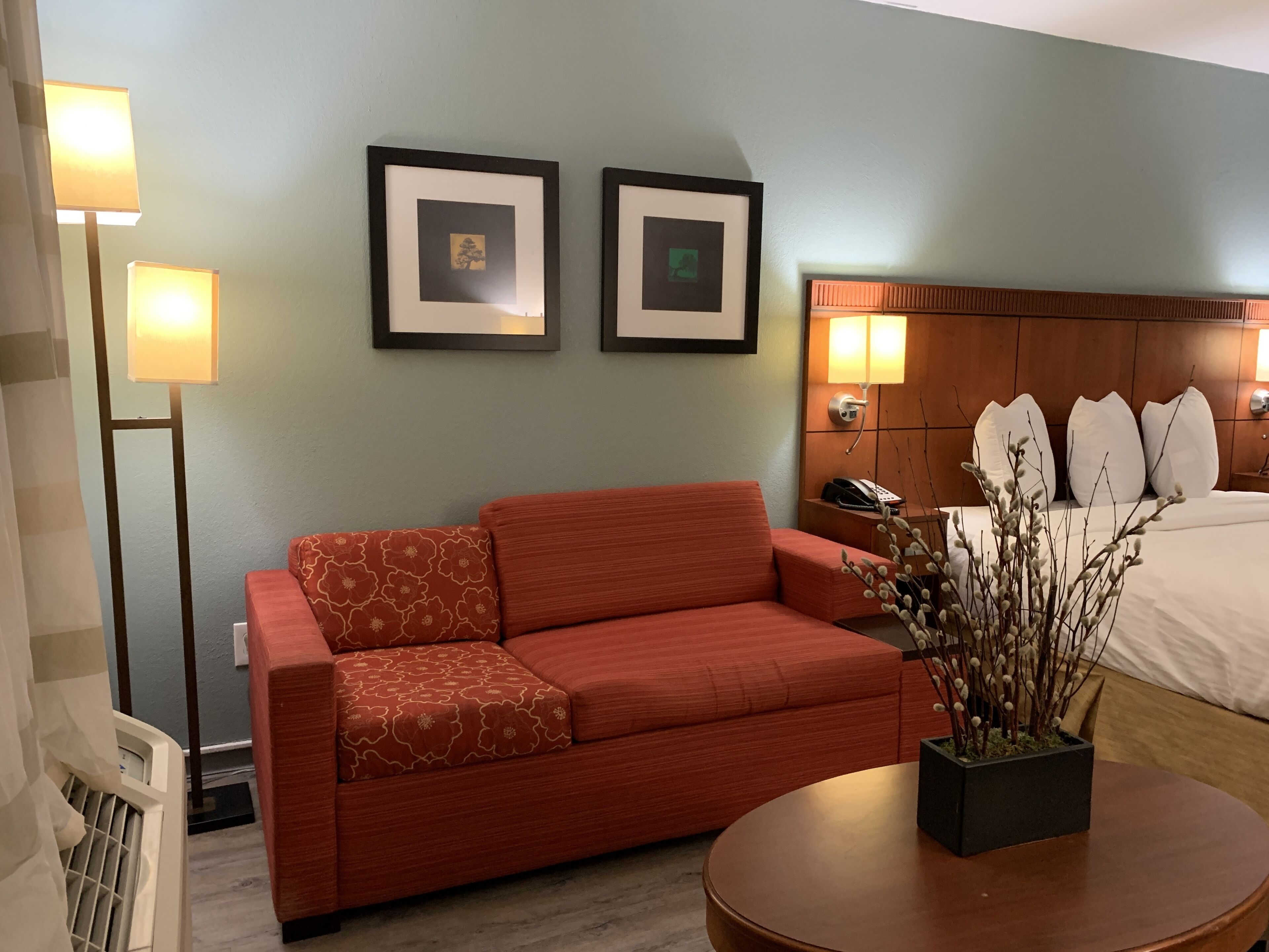 Quarto de hotel com sofá vermelho, mesa de madeira redonda, cama com roupa de cama branca e duas imagens emolduradas na parede.