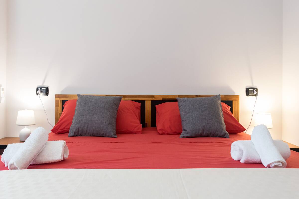 Quarto de hotel com cabeceira de madeira, roupa de cama vermelha, duas almofadas cinzas e duas toalhas brancas.