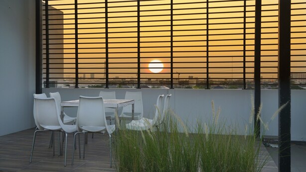 Um terraço com mesa e cadeiras, vista do pôr do sol através de uma janela com persianas, e vegetação ao primeiro plano.