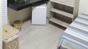 Quarto pequeno e funcional com cama, máquina de lavar roupa branca, guarda-roupa de madeira com prateleiras, espelho e bancada preta com pia.