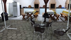 Um pátio externo com mesa de vidro e cadeiras de vime, cercado por fornos de tijolo e uma geladeira.