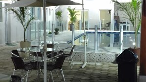 Uma área de pátio ao ar livre com mesa e cadeiras de vidro, rodeada por vasos de plantas e uma piscina.