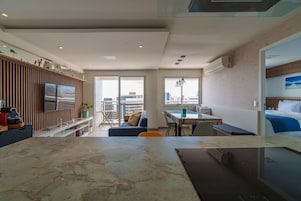 Quarto de hotel moderno com uma janela grande, área de jantar, sala de estar com sofá e cozinha com bancada de mármore.