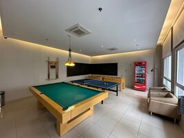 Uma mesa de bilhar, duas mesas de futebol de mesa, uma máquina de vendas vermelha e um sofá numa sala moderna com grandes janelas.