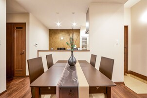 Uma sala de jantar com uma mesa de madeira comprida, cadeiras e um vaso com galhos. Dá para ver uma cozinha ao fundo, com painel e armários de madeira.