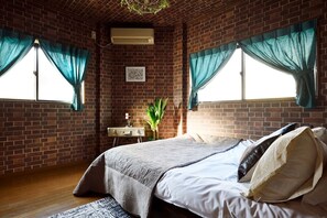 Quarto com parede de tijolos, cama com colcha branca e cinza, duas janelas com cortinas verde-água, uma escrivaninha pequena com abajur e uma planta.