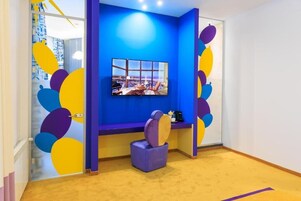 Quarto com uma TV grande numa parede azul, uma cadeira roxa e vista para a cidade.