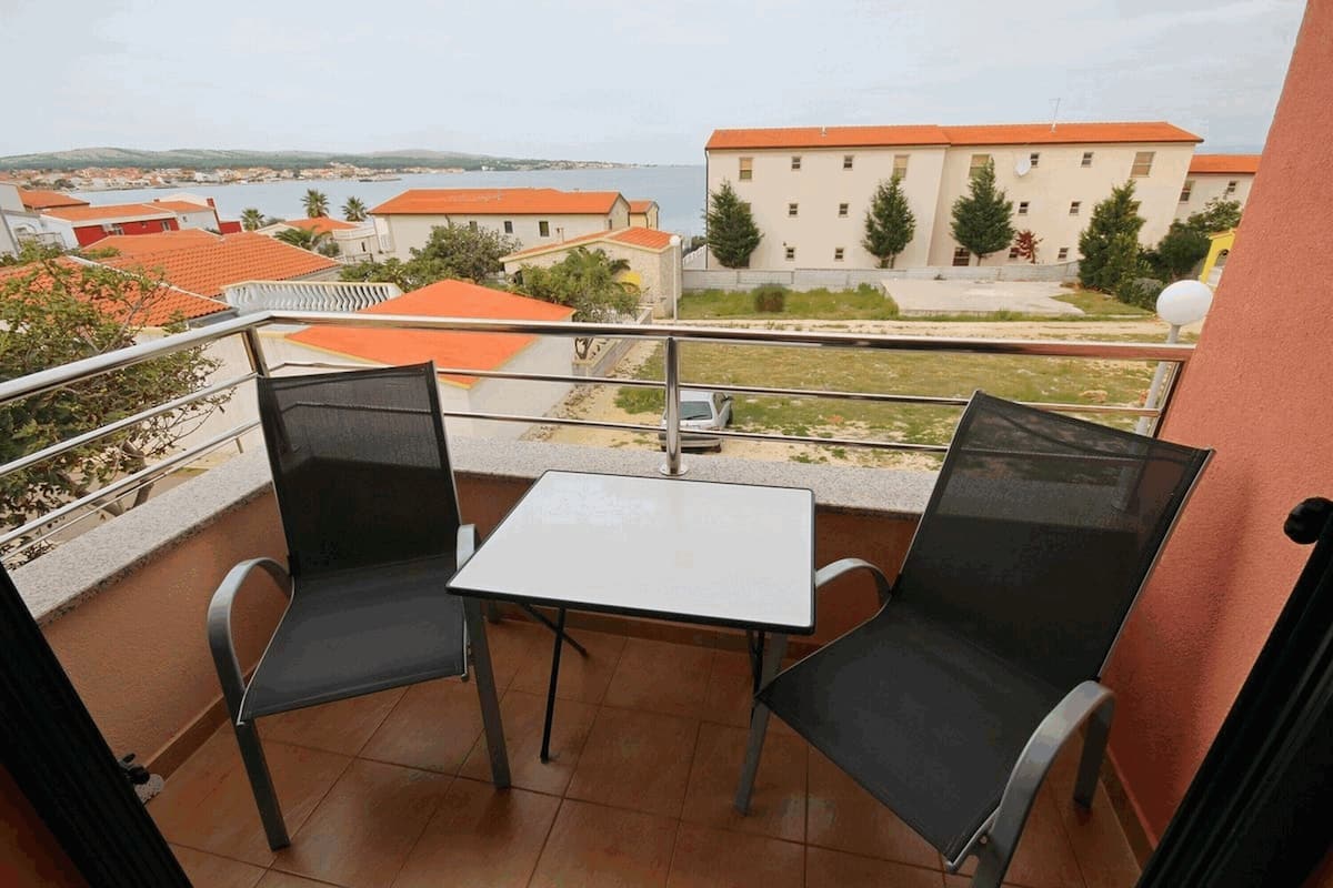 Varanda com duas cadeiras e uma mesa, com vista para a área costeira, edifícios e um corpo de água.