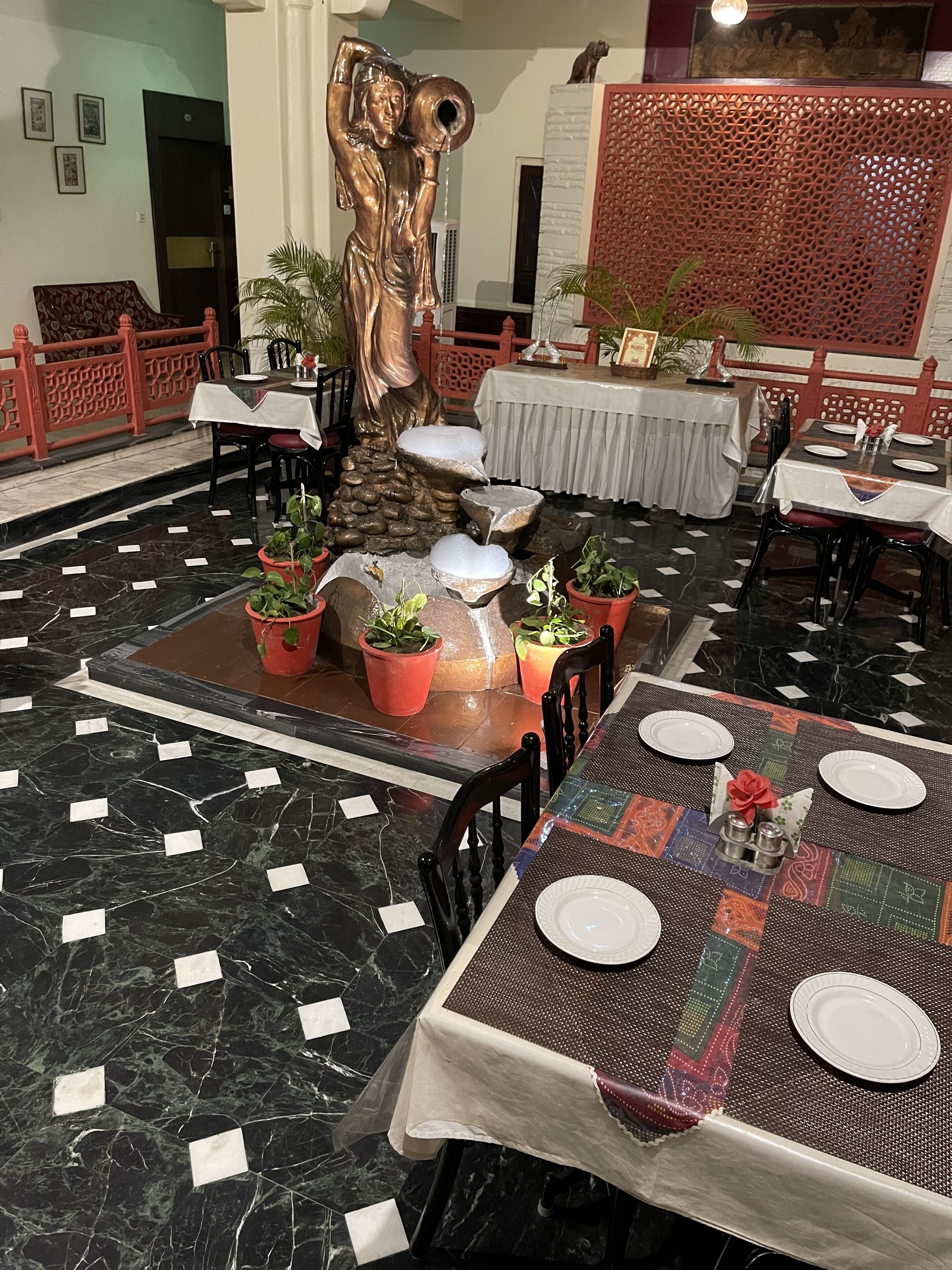 Restaurante com piso de mármore, mesas preparadas com pratos brancos e plantas em vasos.