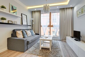 Uma sala de estar moderna com um sofá cinzento, mesa de centro branca e uma TV sobre um armário branco. Há quadros nas prateleiras e uma janela grande com cortinas.